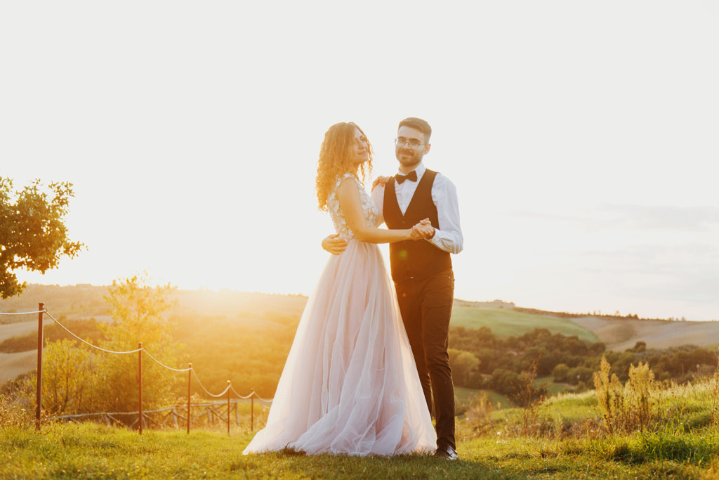 Happy bride and groom standing on field against clear sky during sunset ...
